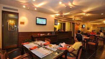Um restaurante com mesas montadas para refeições, uma tela de TV exibindo um jogo esportivo e um balcão de recepção.