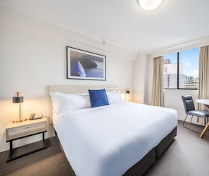 Quarto de hotel com uma cama grande, uma mesa de cabeceira com abajur, uma cadeira e vista para a cidade pela janela.