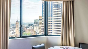 Quarto de hotel com uma janela ampla que oferece vista para a cidade, uma mesa redonda com uma revista e uma cadeira.