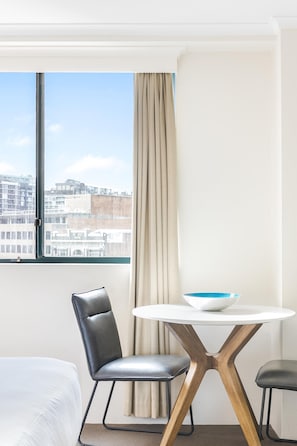 Quarto de hotel com cama, cadeira, mesa com tigela, janela com vista para prédios e cortina.
