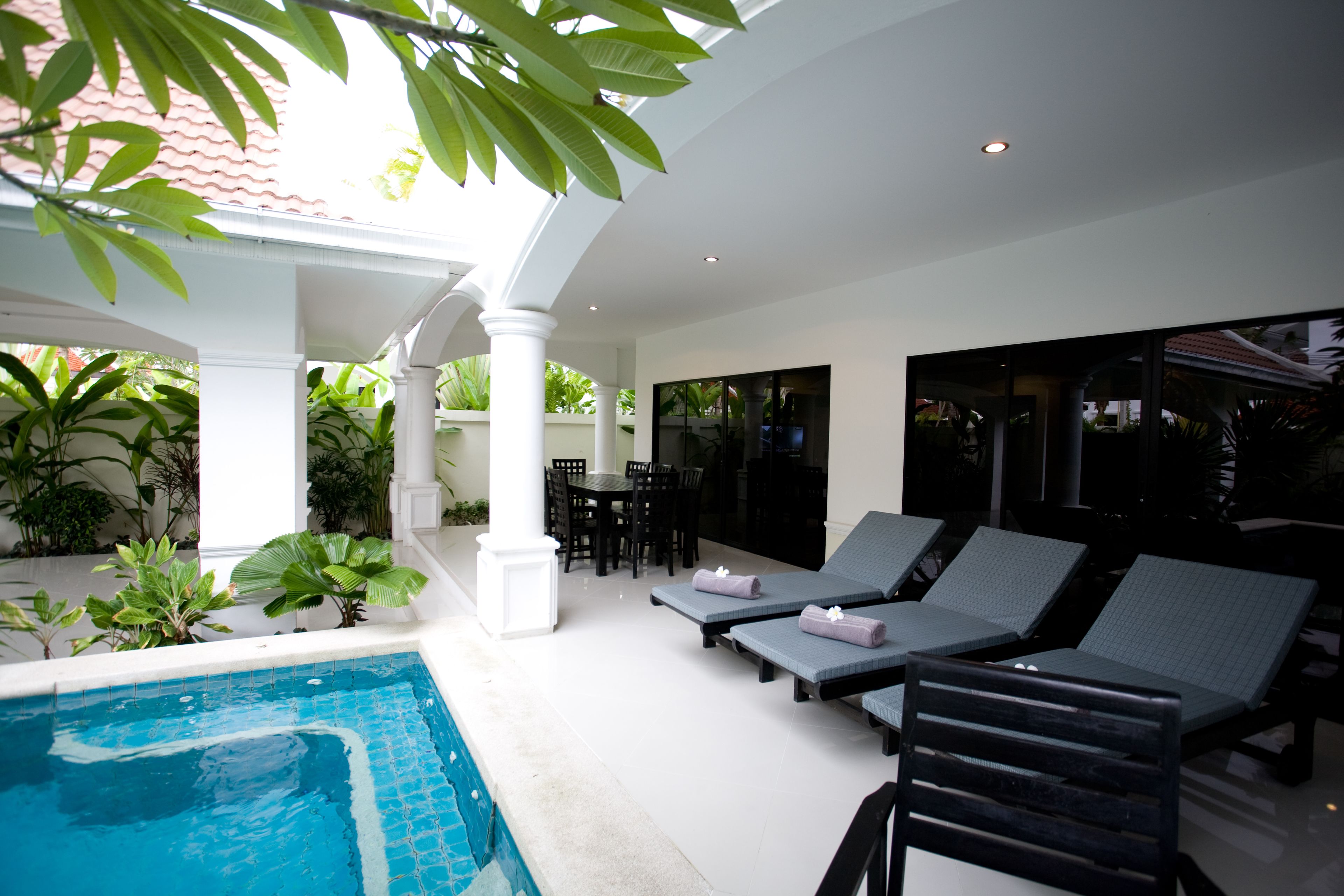 Uma área à beira da piscina com espreguiçadeiras, uma mesa e cadeiras, cercada por plantas e vista desobstruída da piscina.