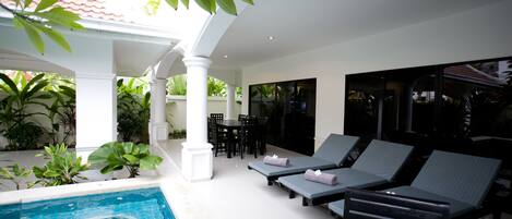 Uma área à beira da piscina com espreguiçadeiras, uma mesa e cadeiras, cercada por plantas e vista desobstruída da piscina.