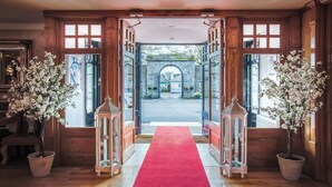 Entrada de hotel com tapete vermelho, portas de madeira e flores brancas em vasos.