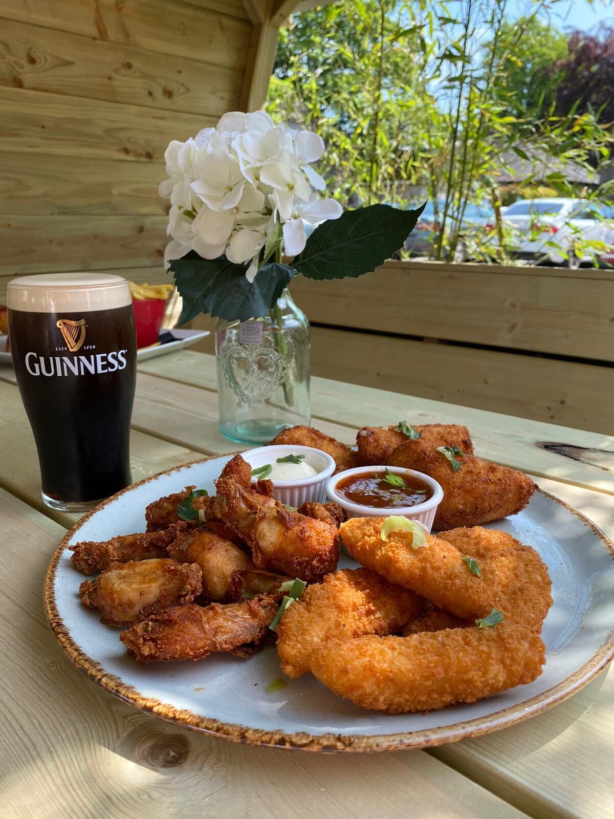 Um prato de coxas de frango crocantes com molhos para acompanhar, um copo de cerveja Guinness e um vaso com flores brancas sobre uma mesa de madeira.