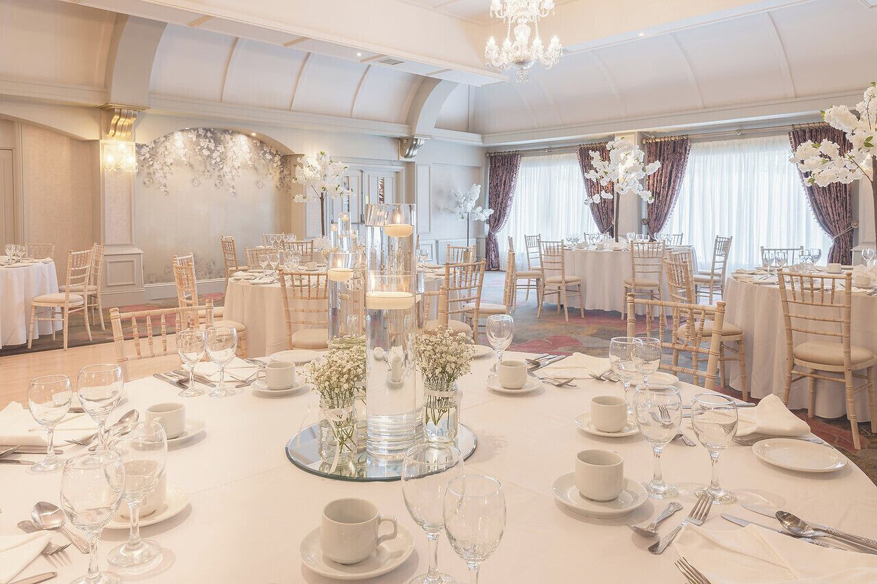 Salão de banquetes com mesas redondas preparadas para refeições, com toalhas brancas, copos e centros de mesa florais.