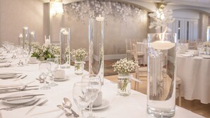 Salão de festas com longas mesas preparadas para um evento formal, com castiçais de vidro transparente e arranjos florais.