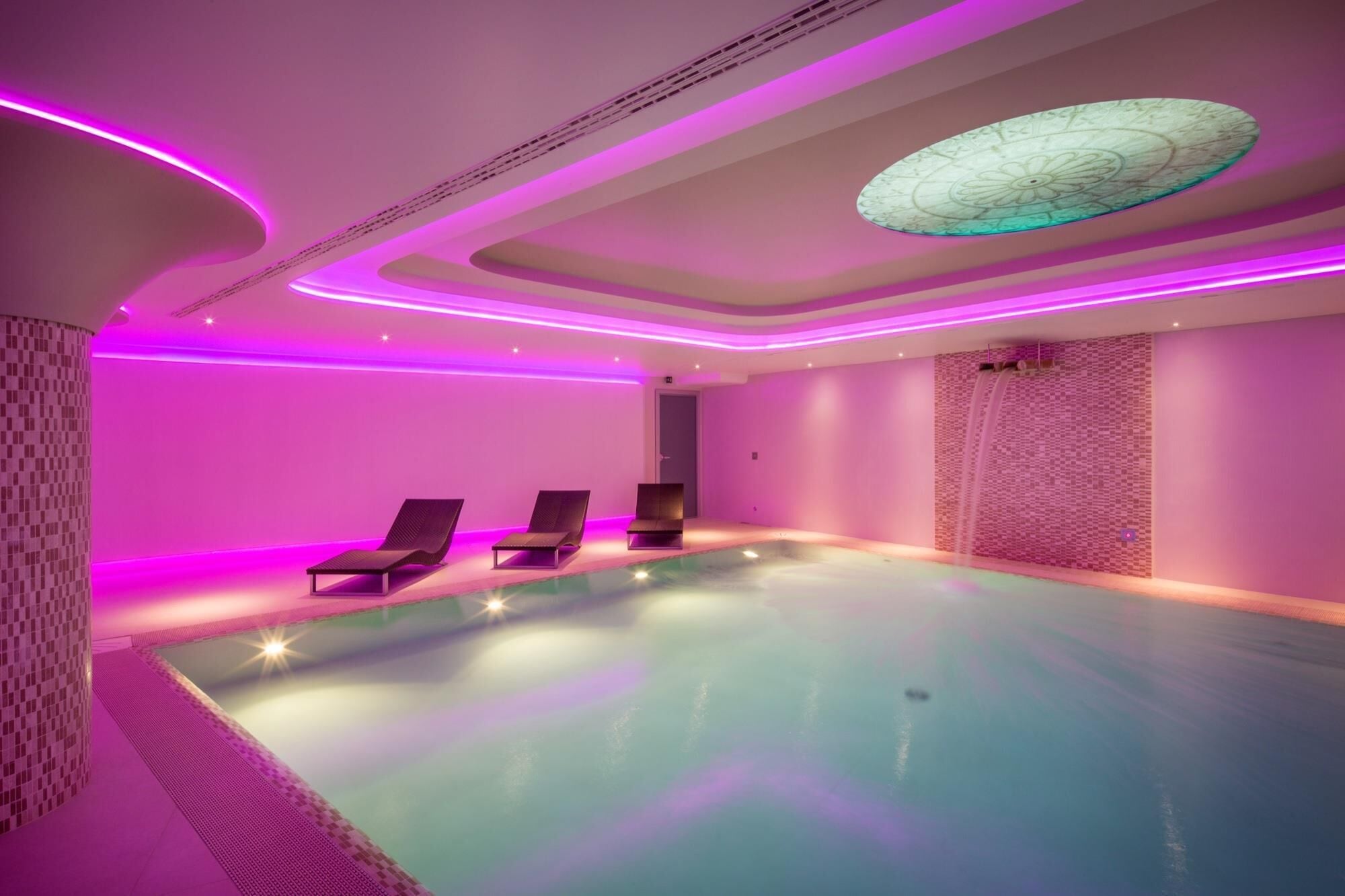 Piscina interior moderna com iluminação rosa suave, três espreguiçadeiras e teto decorativo.