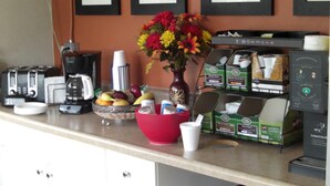 Uma bancada de cozinha com cafeteira, torradeira e uma tigela de frutas. Também há caixas de café e um vaso com flores.