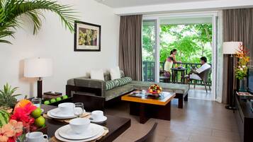 Quarto de hotel com área de jantar, sofá, varanda com vista para árvores e televisão.