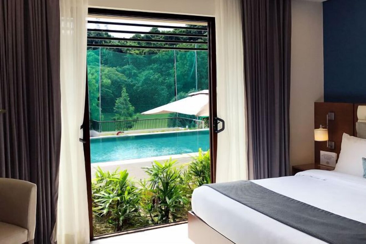 Quarto de hotel com uma janela grande com vista para uma piscina e vegetação exuberante.