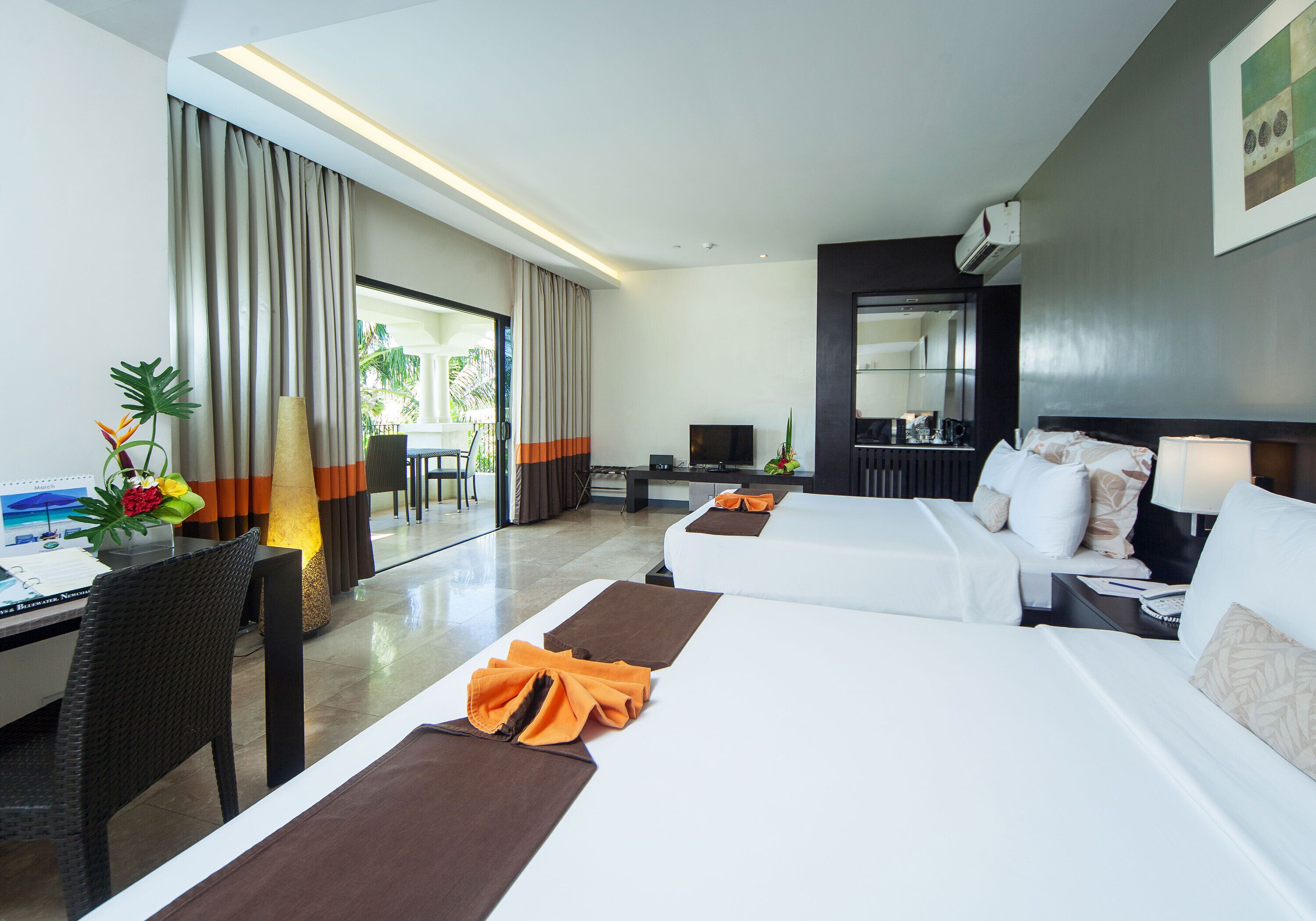 Quarto de hotel com duas camas, uma escrivaninha, uma cadeira, uma televisão e uma janela grande com cortinas.