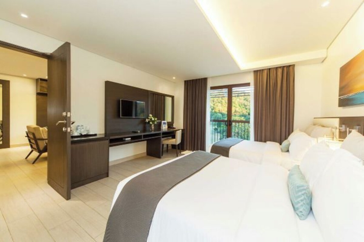 Quarto de hotel moderno com uma cama grande, uma mesa de trabalho com televisão de tela plana, um espaço de estar e uma varanda com cortinas.