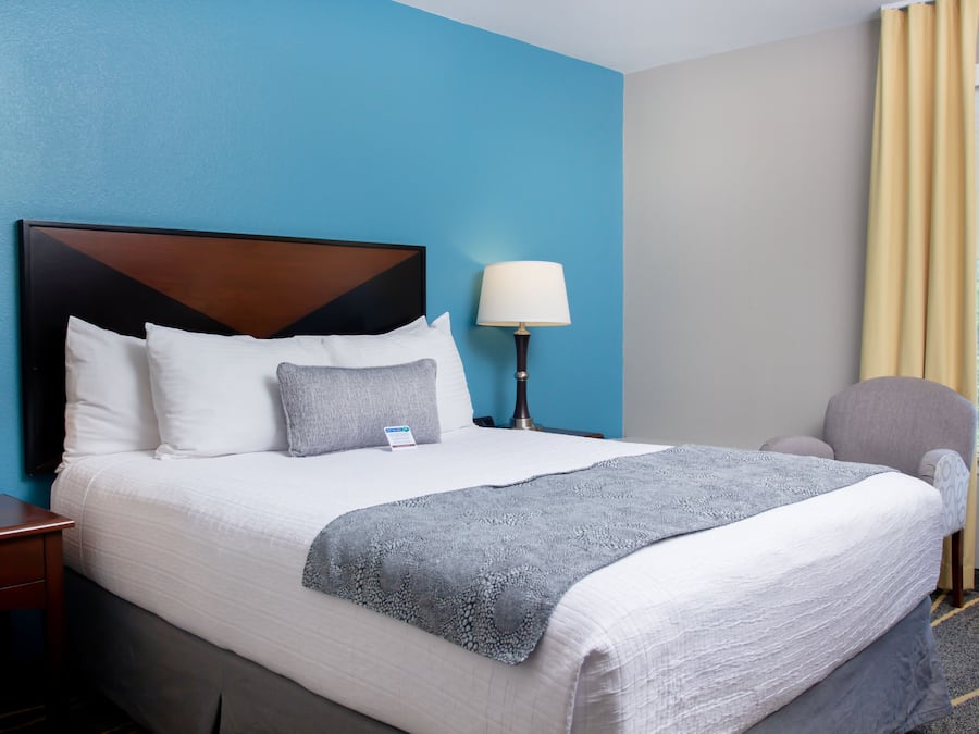 Quarto de hotel com uma cama grande, duas luminárias de cabeceira, uma cadeira e uma janela com cortinas.