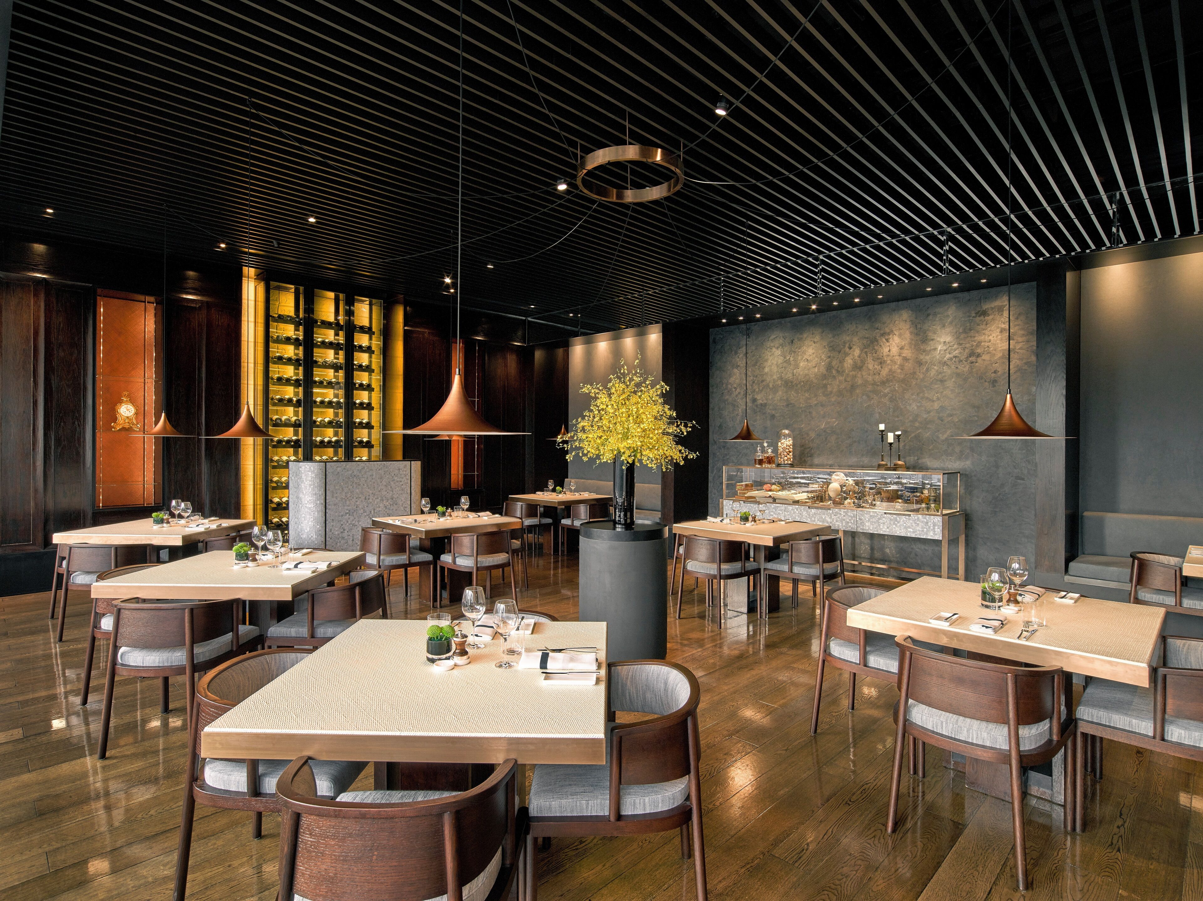 Interior moderno de restaurante com várias mesas e cadeiras, um aparador de vinhos e um vaso grande com flores.