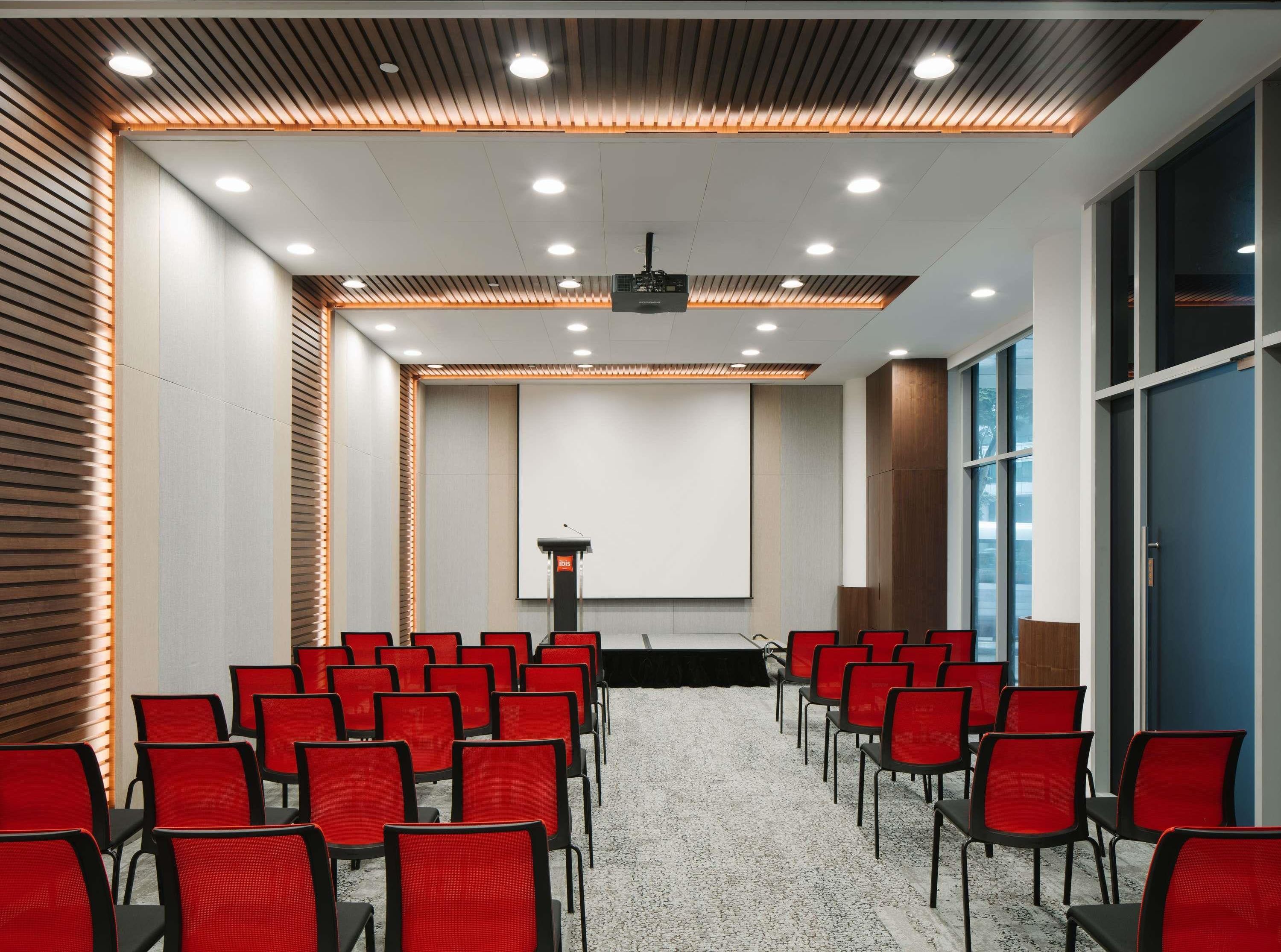 Uma sala de conferências com fileiras de cadeiras vermelhas, um púlpito, uma tela de projetor e uma grande janela.
