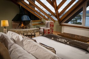 Um quarto acolhedor com lareira de pedra, uma poltrona confortável, uma cama com roupa de cama branca e vista para o exterior através de uma grande janela.