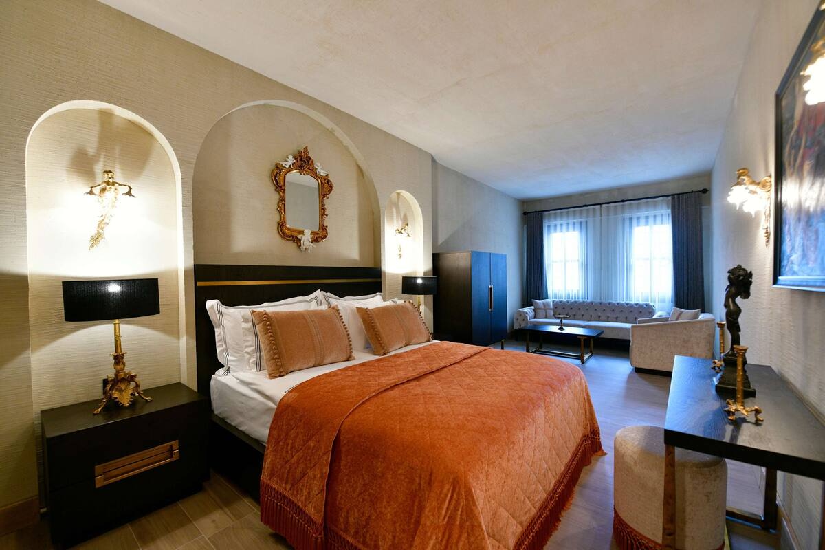 Quarto de hotel com uma cama grande, área de estar com sofá e poltronas, escrivaninha com luminária e um espelho grande na parede.