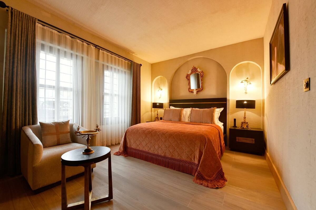Quarto de hotel com cama, sofá, mesa e duas luminárias fixadas na parede.