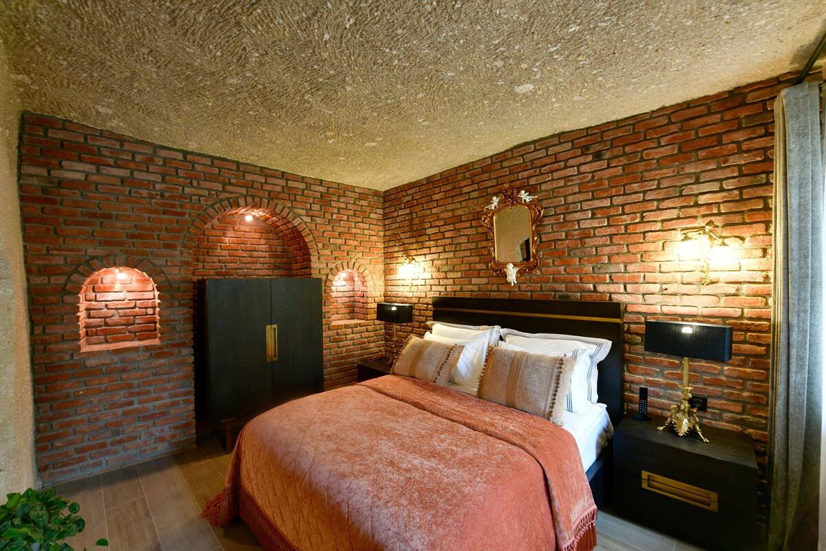 Quarto com parede de tijolos, cama com cabeceira, mesa de cabeceira com abajur e espelho na parede.