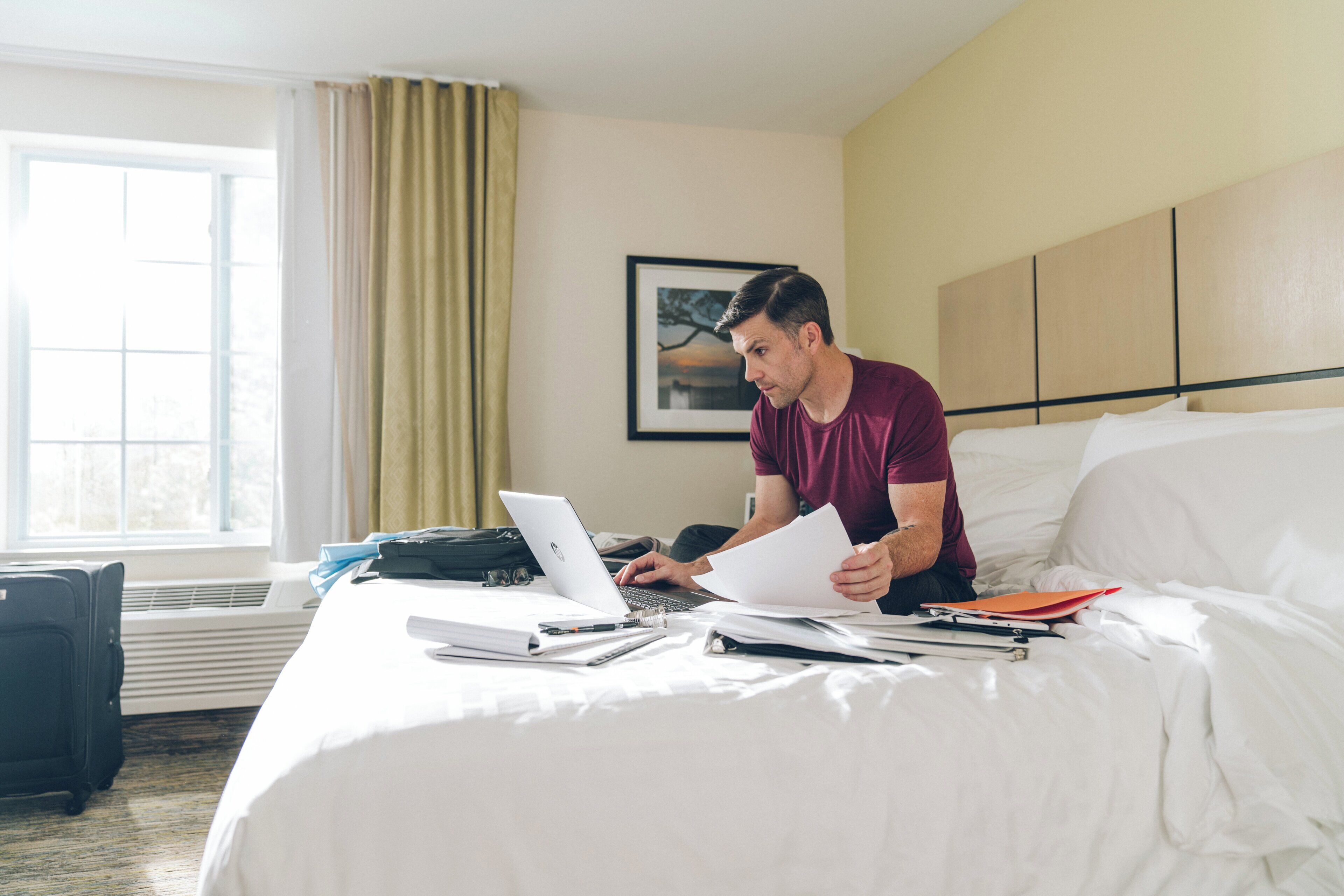 Quarto de hotel com cama, laptop, papéis e mala.