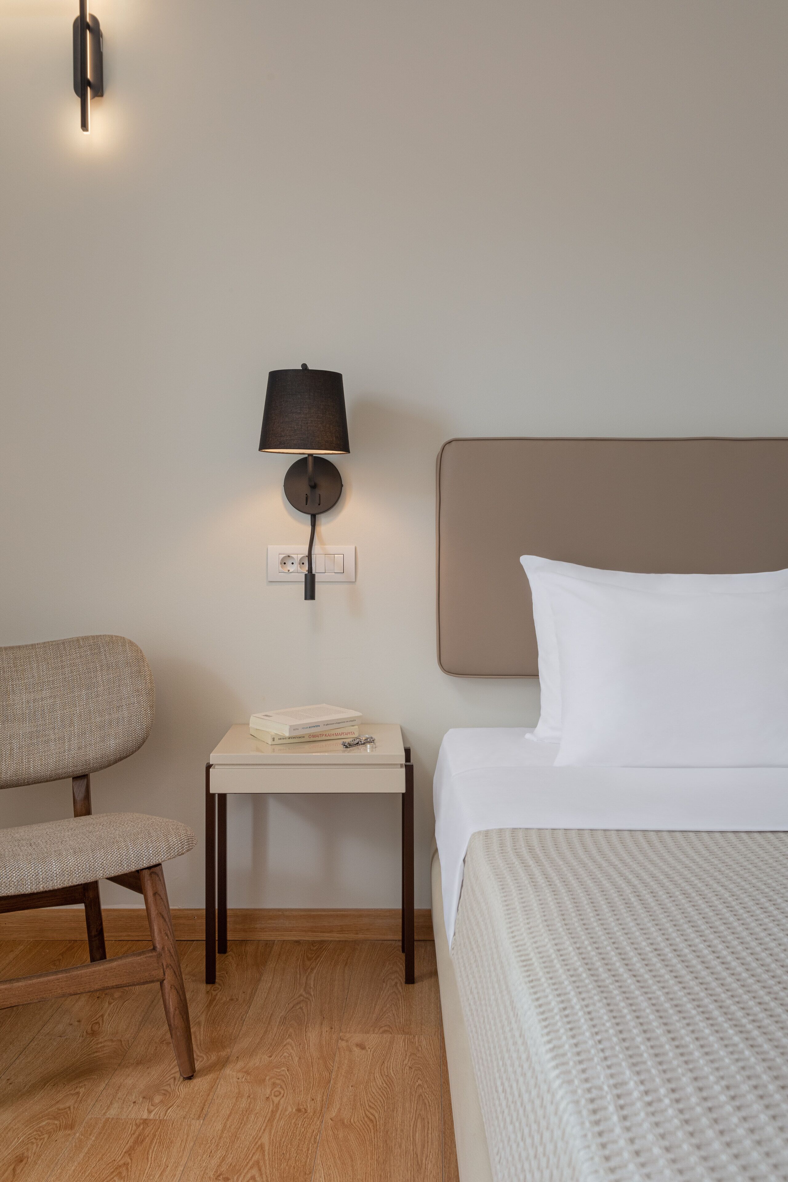 Quarto de hotel com cama, uma cadeira, uma pequena mesa com um livro, uma luminária na parede e uma tomada.