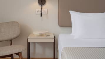 Quarto de hotel com cama, uma cadeira, uma pequena mesa com um livro, uma luminária na parede e uma tomada.