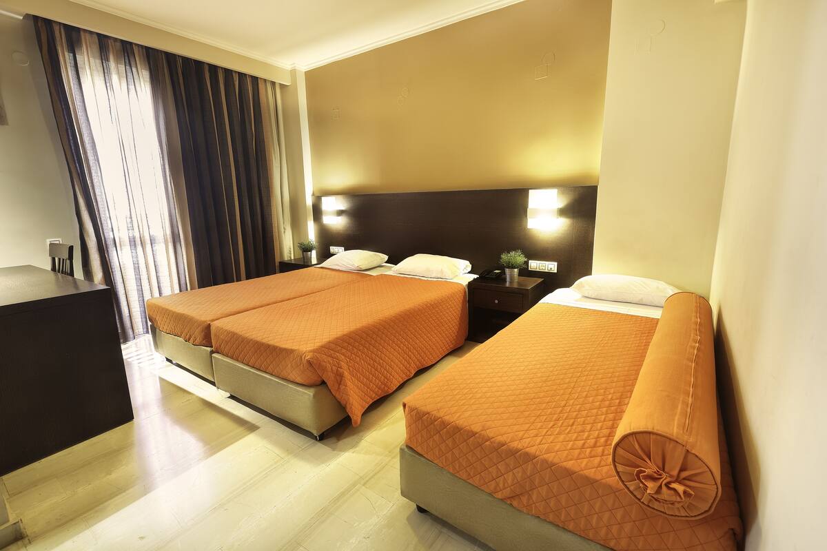 Quarto de hotel com duas camas, uma escrivaninha, uma mesa de cabeceira e uma janela grande com cortinas.