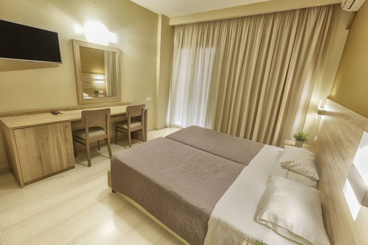 Quarto de hotel com cama, escrivaninha, cadeira, televisão e janela com cortinas.