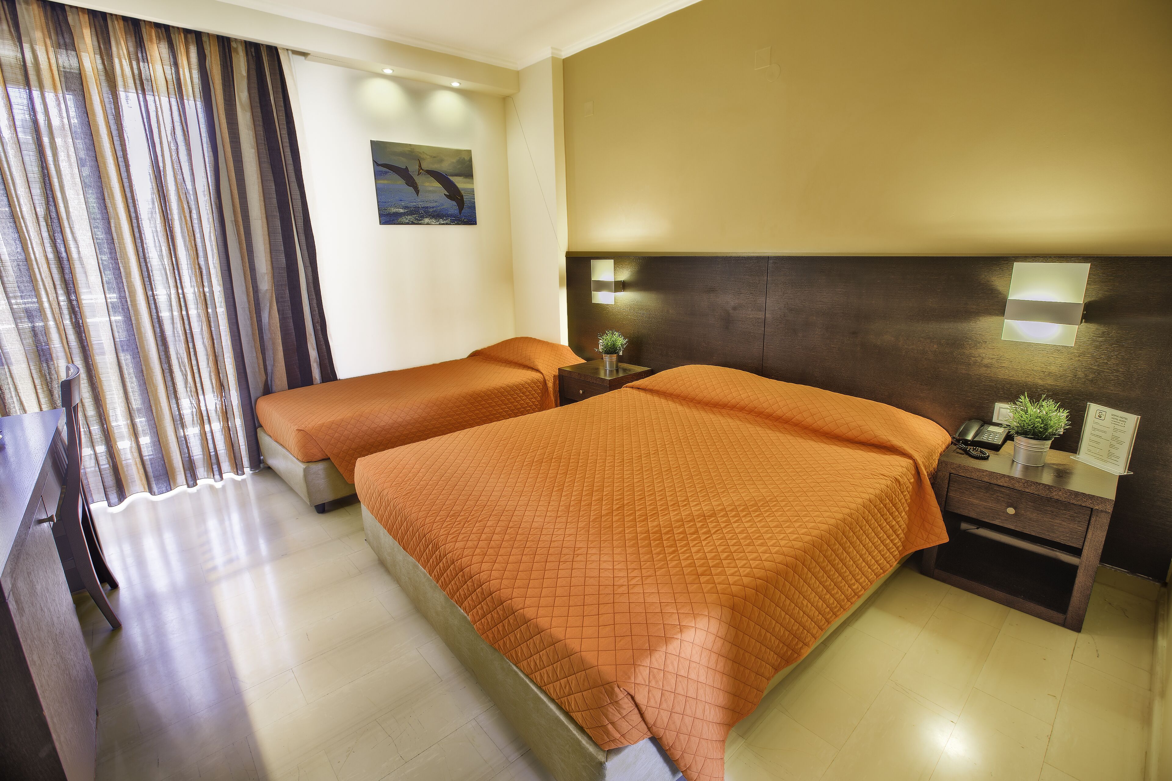 Quarto de hotel com duas camas, uma escrivaninha, uma cadeira, mesa de cabeceira com abajur e um quadro na parede.