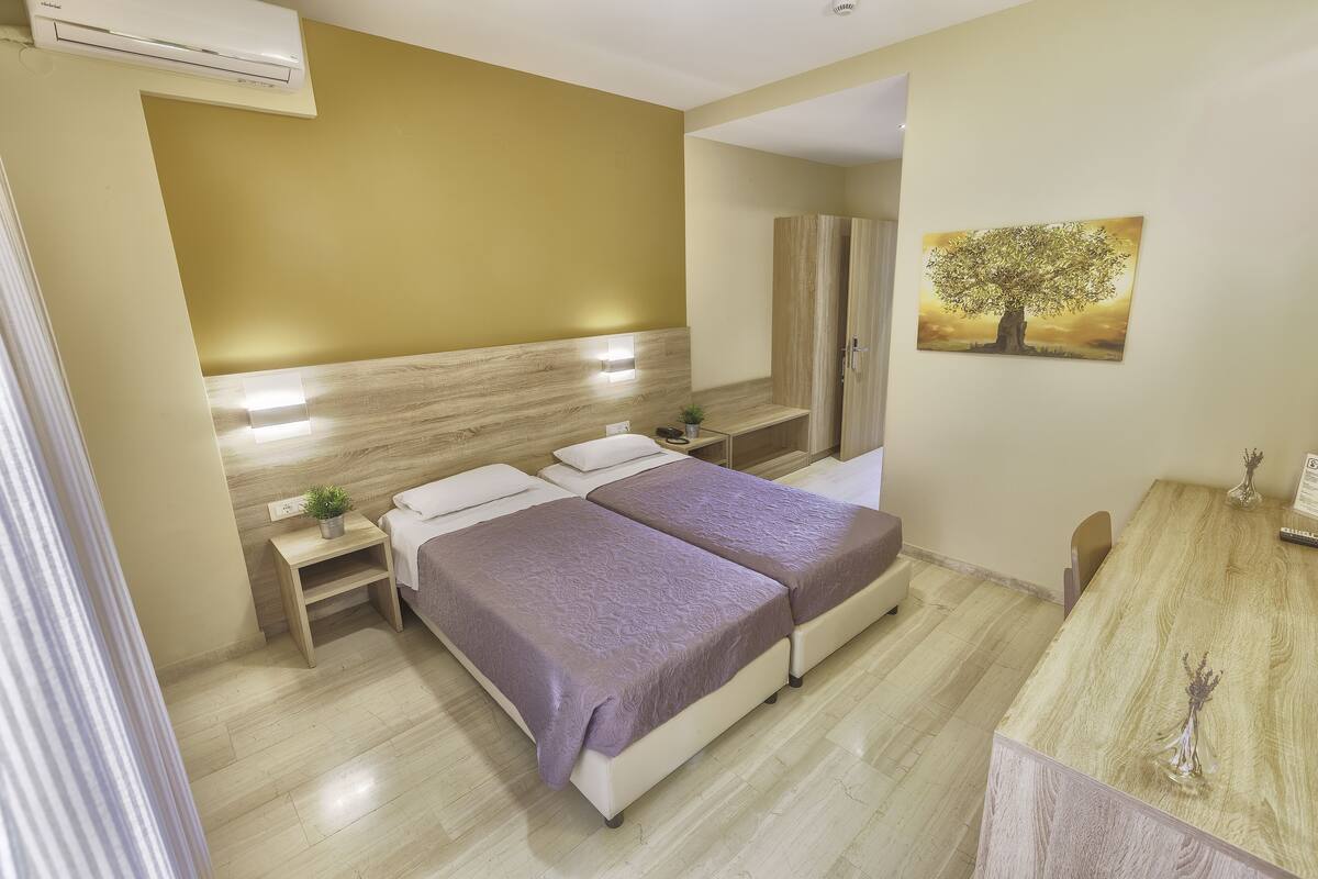 Quarto de hotel com duas camas, cabeceira de madeira, mesa de cabeceira com uma planta, pintura de uma árvore e escrivaninha de madeira com um vaso.
