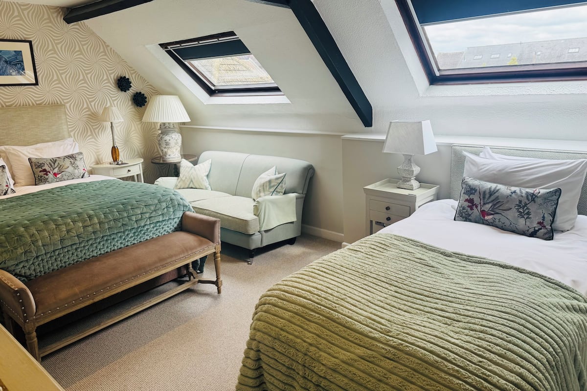 Quarto com teto estilo loft, cama com colcha verde, sofá com manta clara, um banco e uma mesa de cabeceira com abajur.