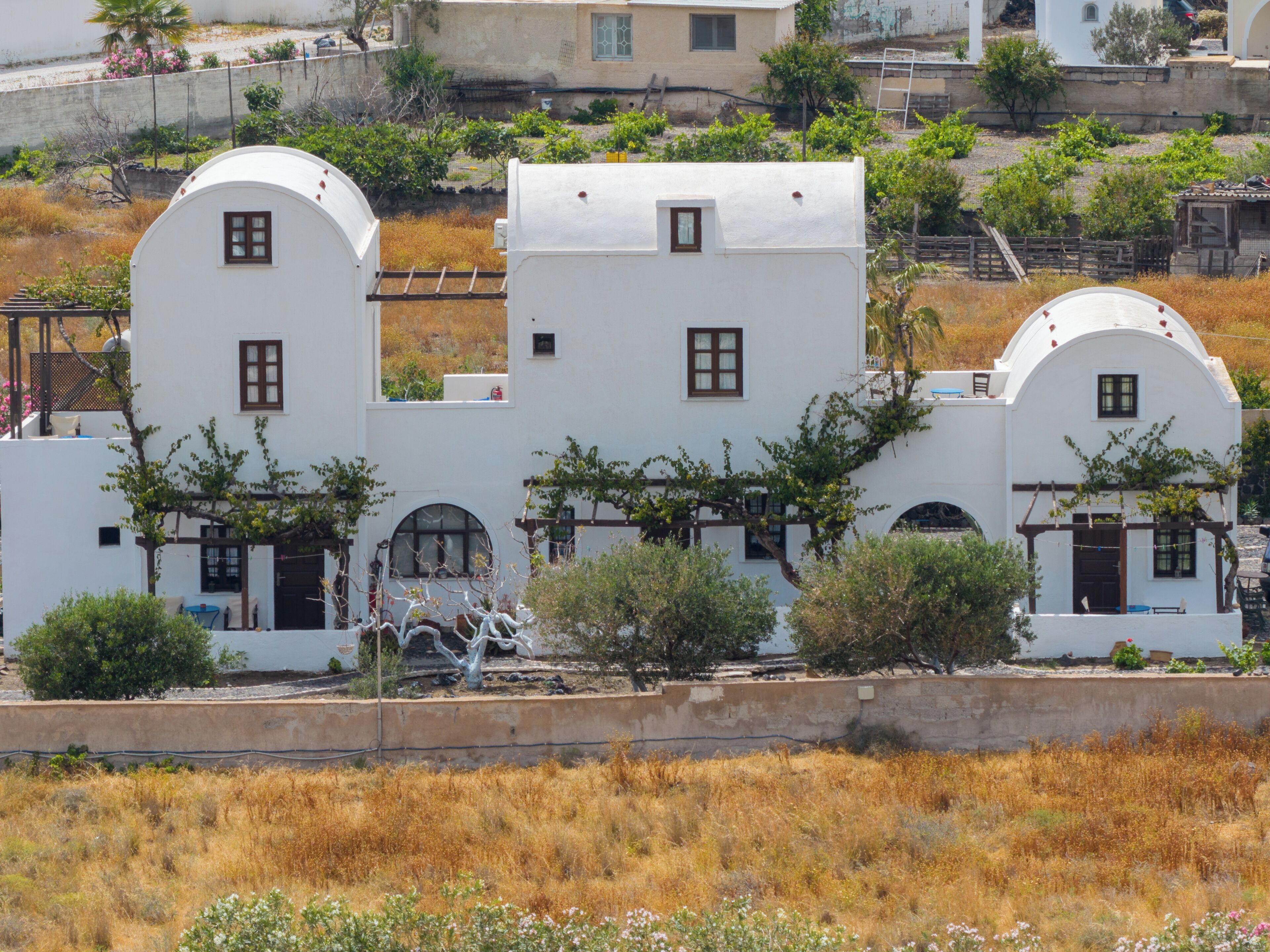 Um edifício branco com janelas em arco e telhado plano, rodeado por relva seca e vegetação escassa.