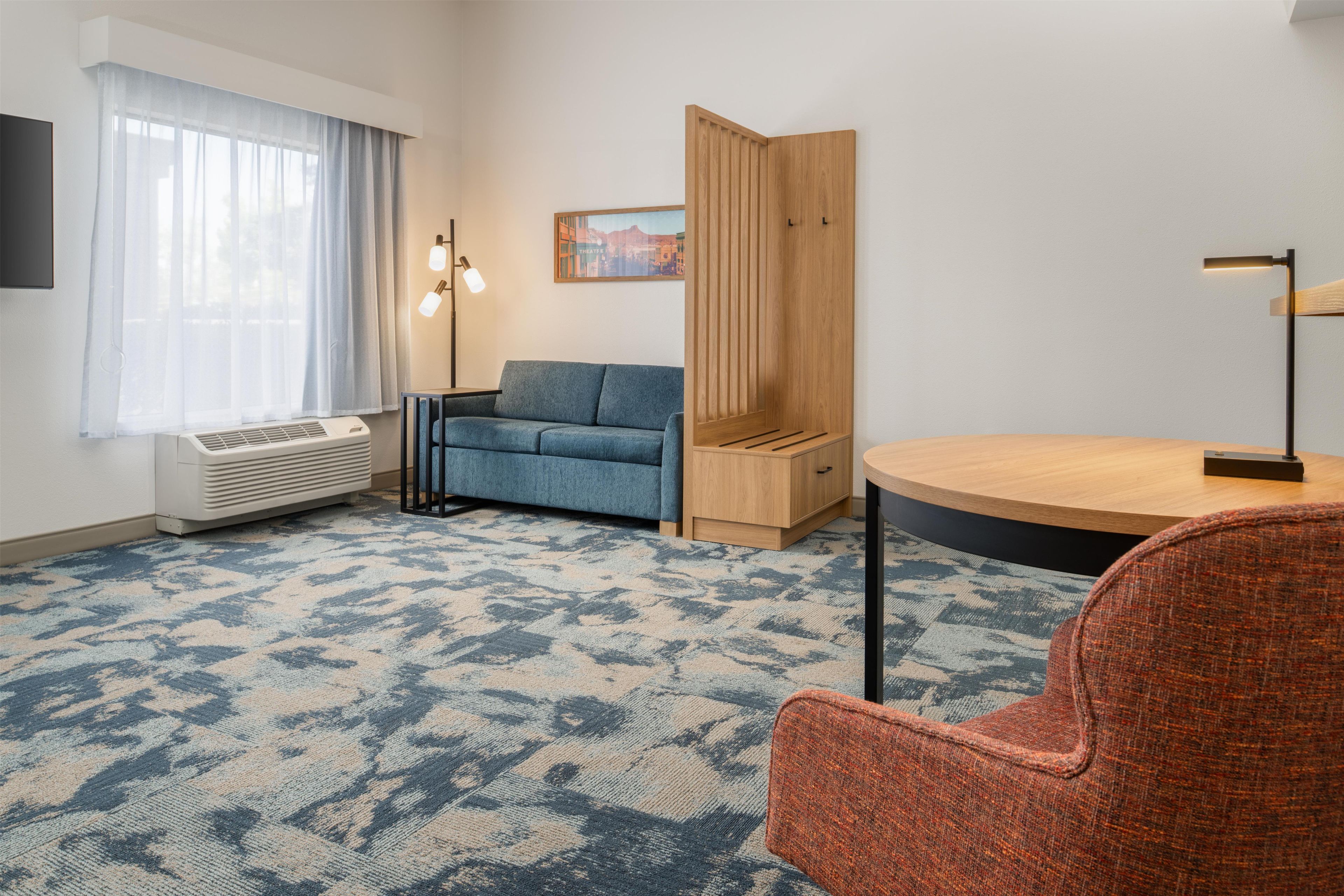 Quarto de hotel com um sofá azul, um armário de madeira, uma mesa redonda e um abajur.