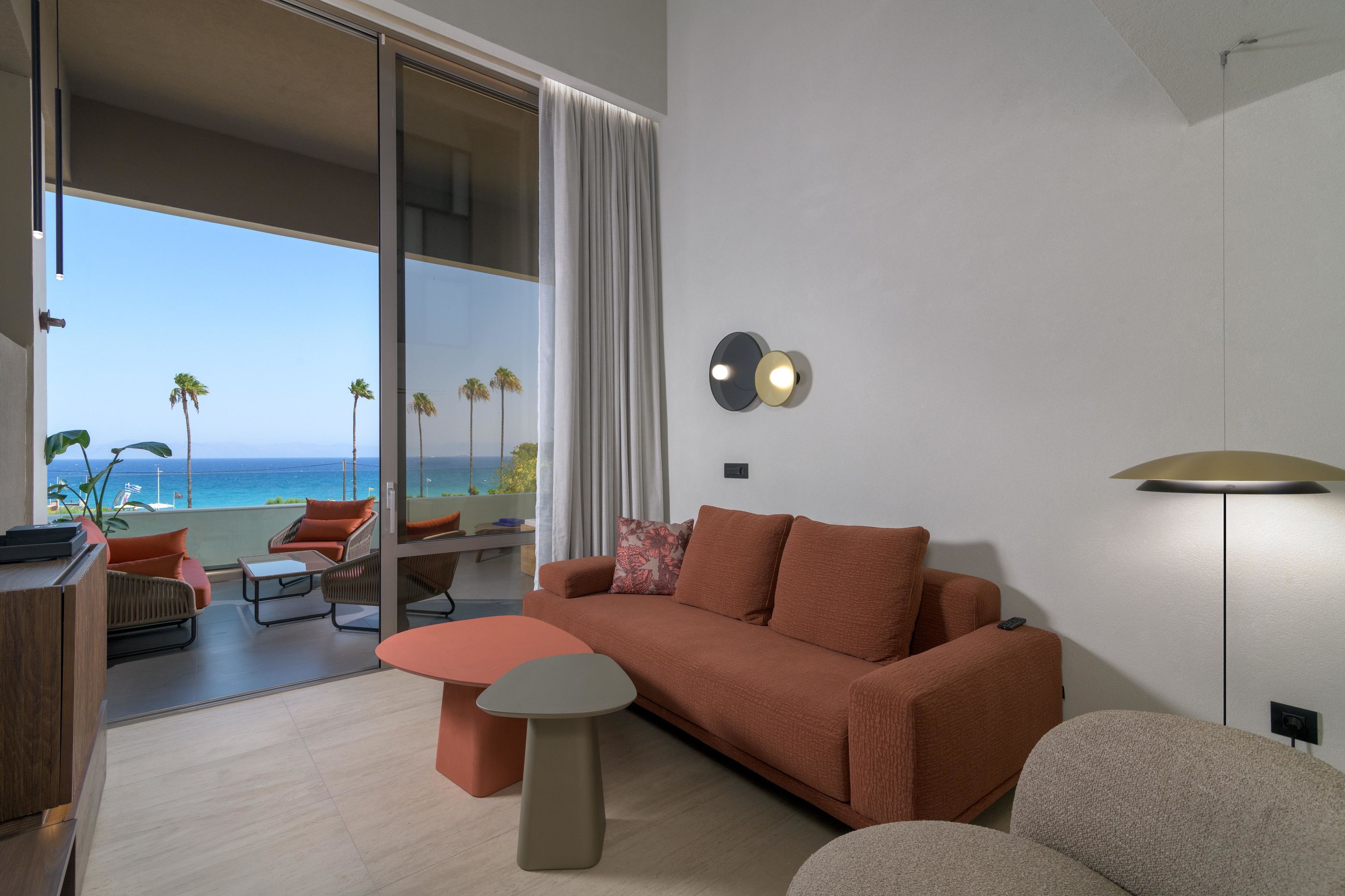 Uma sala de estar moderna com uma grande janela com vista para uma praia, um sofá, uma mesa de centro e um abajur.