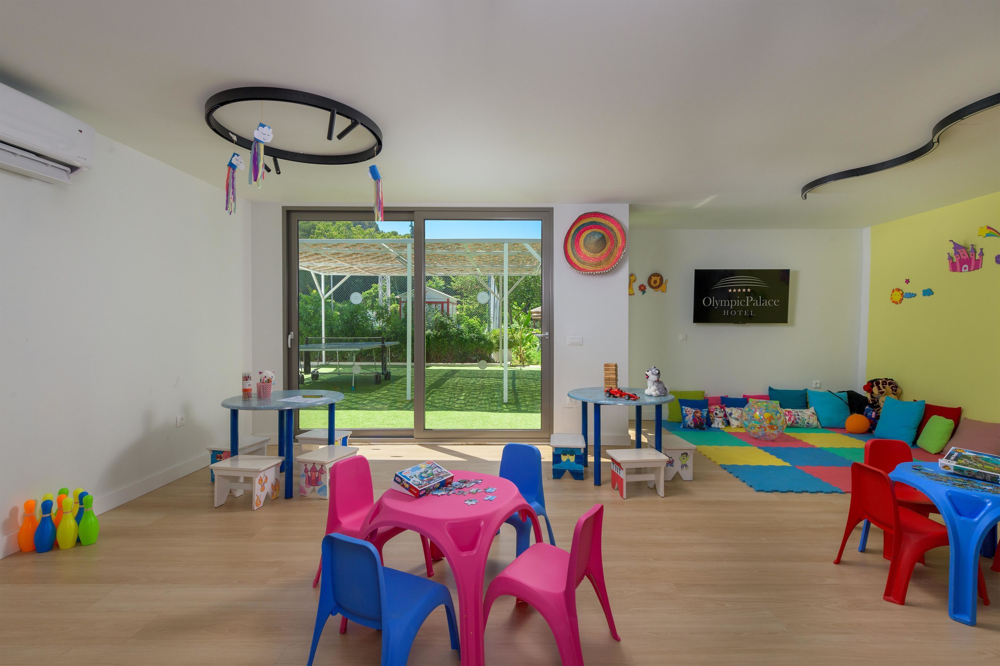 Uma sala de brinquedos infantil com mobiliário e brinquedos coloridos, uma janela grande com vista para o jardim, e uma placa do Olympic Palace Hotel.