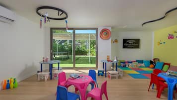 Uma sala de brinquedos infantil com mobiliário e brinquedos coloridos, uma janela grande com vista para o jardim, e uma placa do Olympic Palace Hotel.