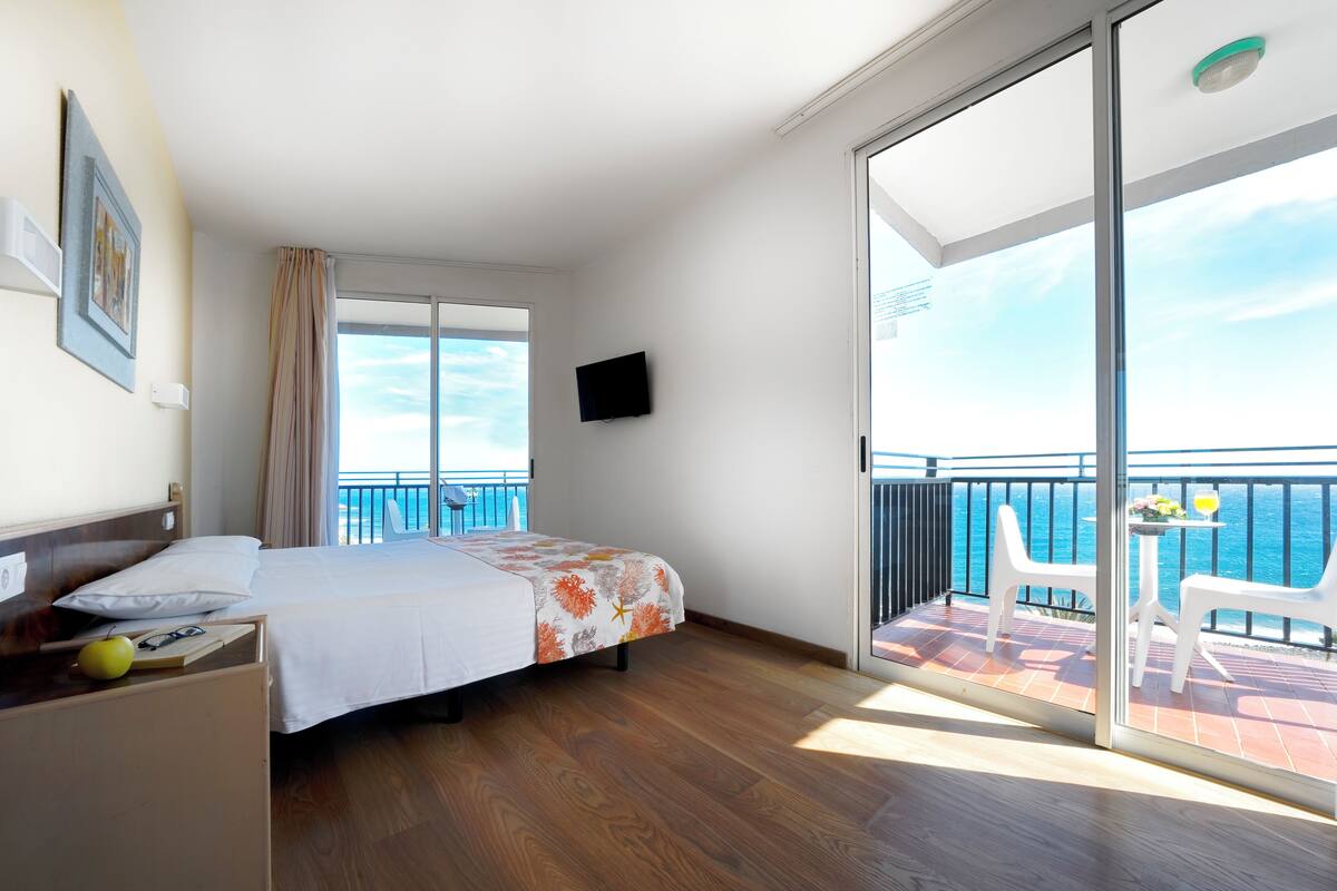 Quarto de hotel com cama, secretária, cadeira, televisão e varanda com vista para o mar.