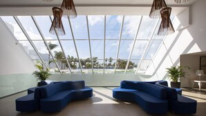 Uma área de estar moderna com um sofá azul curvo, janelas amplas e vista para palmeiras.
