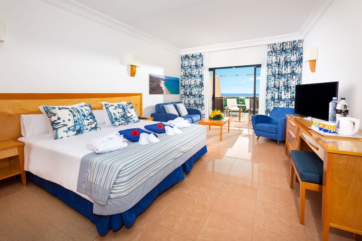 Quarto de hotel com cama, escrivaninha, cadeira, televisão e vista para o mar.