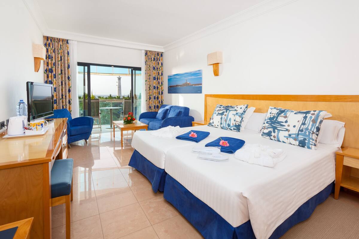 Quarto de hotel com duas camas, uma escrivaninha com televisão, uma cadeira e vista para o exterior.