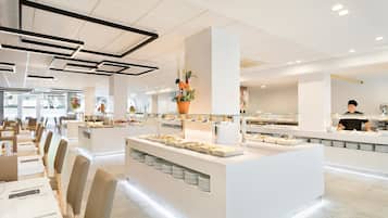 Uma área de refeições moderna com mesas e cadeiras brancas, um buffet central e uma grande planta em vaso.