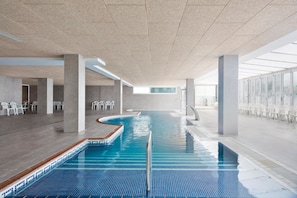 Piscina interior com água azul cristalina, rodeada por cadeiras brancas e chão ladrilhado.