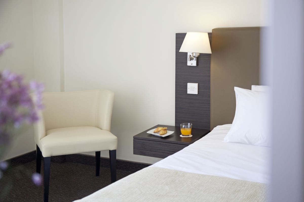Quarto de hotel com cama, uma cadeira, mesa de cabeceira com abajur e uma pequena mesa com um prato de croissants e um copo de sumo de laranja.
