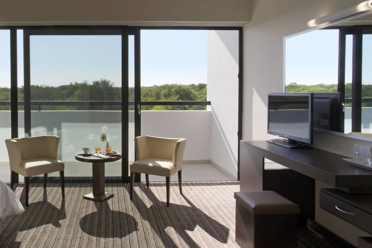 Quarto de hotel com uma cama, duas poltronas, uma pequena mesa com uma flor, uma televisão e uma varanda com vista para o verde.