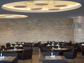 Restaurante com várias mesas e cadeiras, parede de pedra e luminárias circulares no teto.