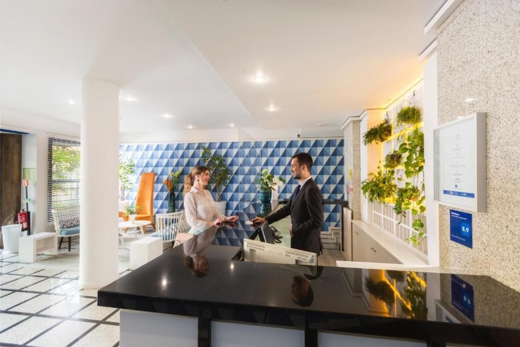 Uma área de recepção com balcão de check-in, parede geométrica azul e plantas dentro de casa.