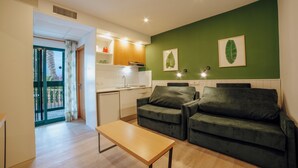 Uma sala de estar moderna com uma mesa de madeira, dois sofás verdes, uma área de cozinha com armários brancos e uma parede verde com arte de folhas.