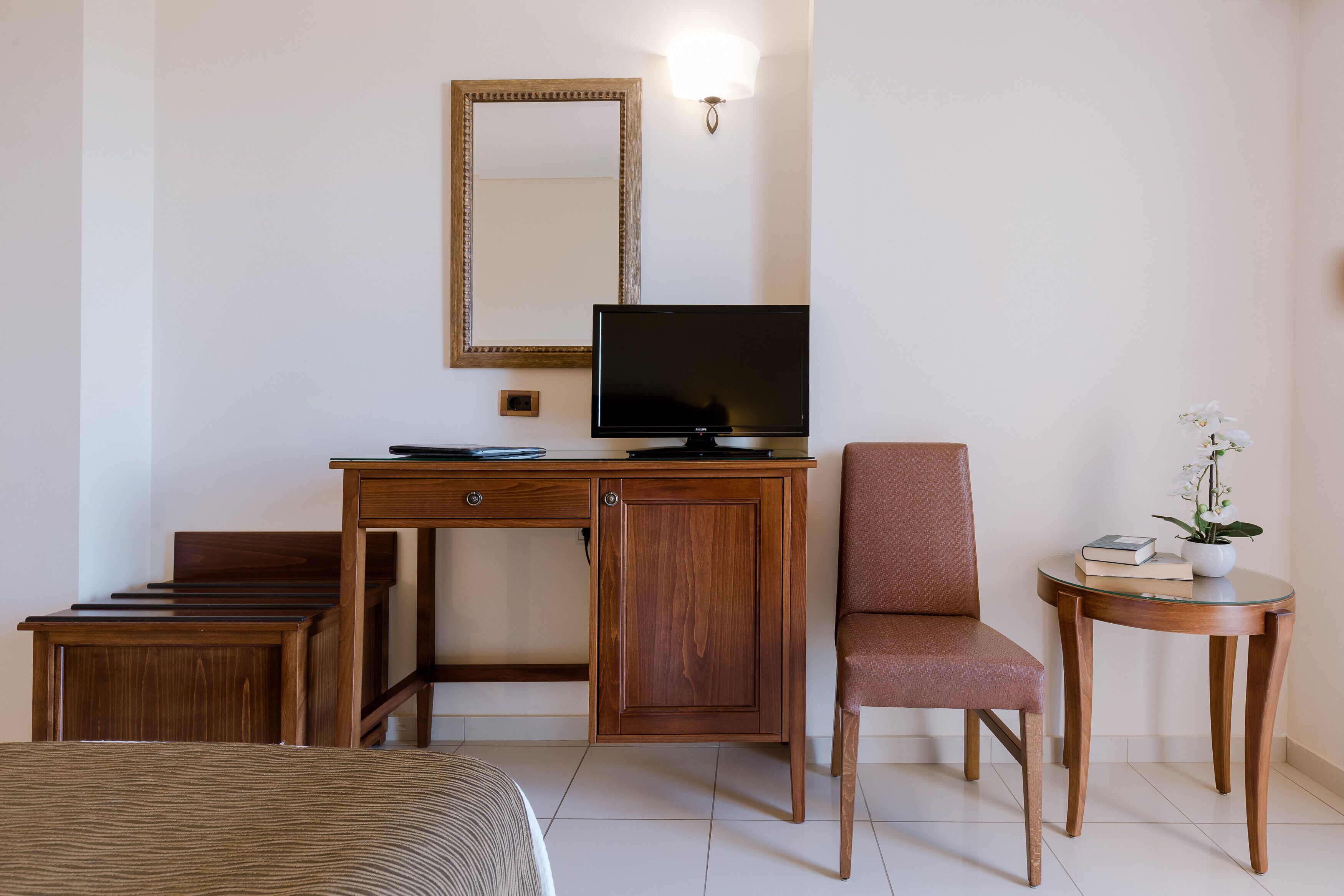 Quarto de hotel com uma escrivaninha de madeira, televisão, cadeira, mesa pequena com uma planta e uma cama.