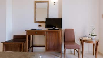 Quarto de hotel com uma escrivaninha de madeira, televisão, cadeira, mesa pequena com uma planta e uma cama.