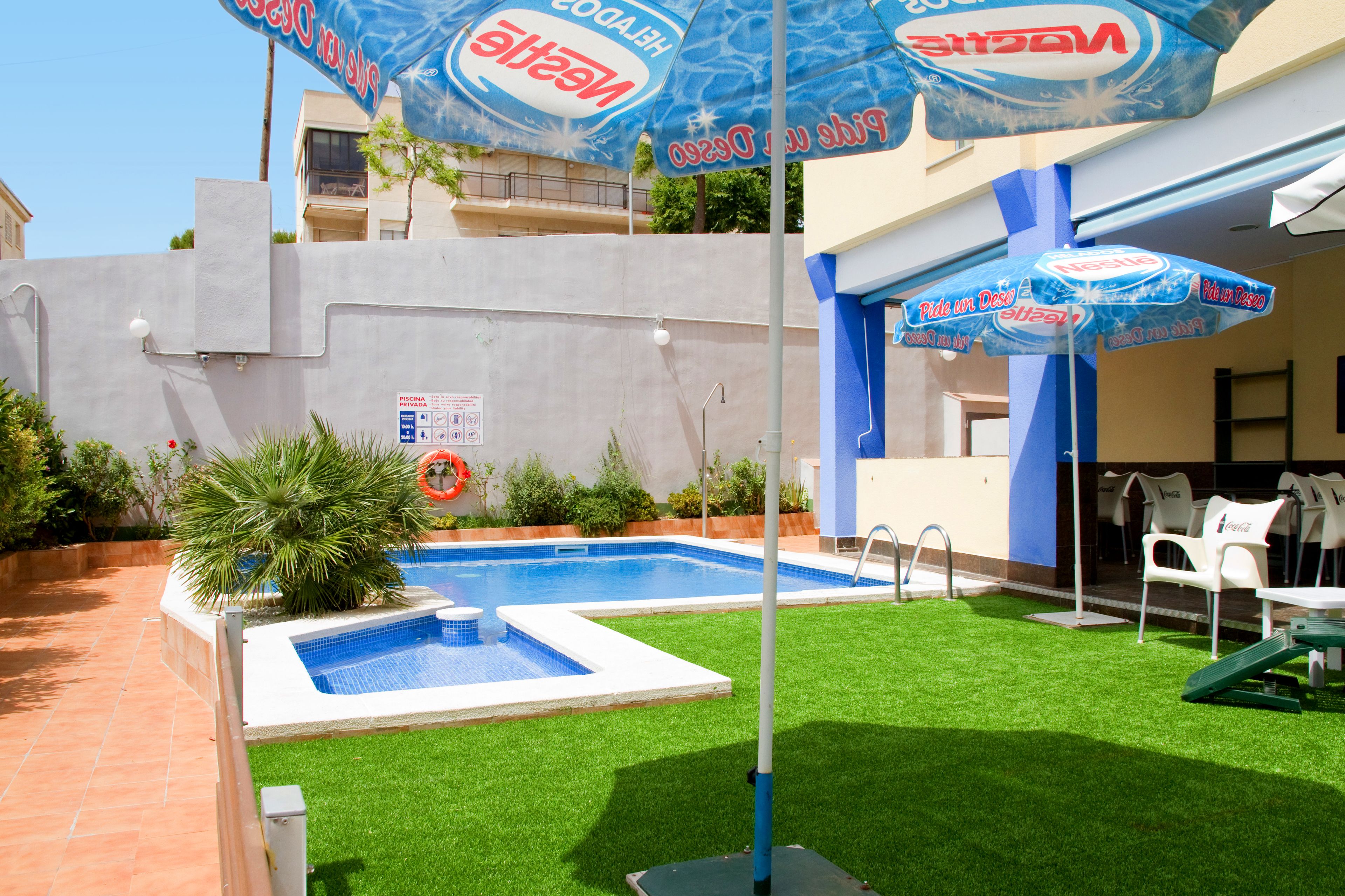 Piscina com grama sintética, um tobogã e guarda-sóis com publicidade de uma marca de água.